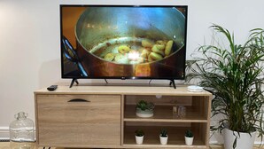 Uma televisão sobre um móvel de madeira com um controle remoto, uma pequena planta e um pote de vidro.