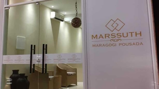 Um interior moderno com portas de vidro, um enfeite esférico pendurado e uma placa com a inscrição "MARSSUTH MARAGOGI POUSADA".