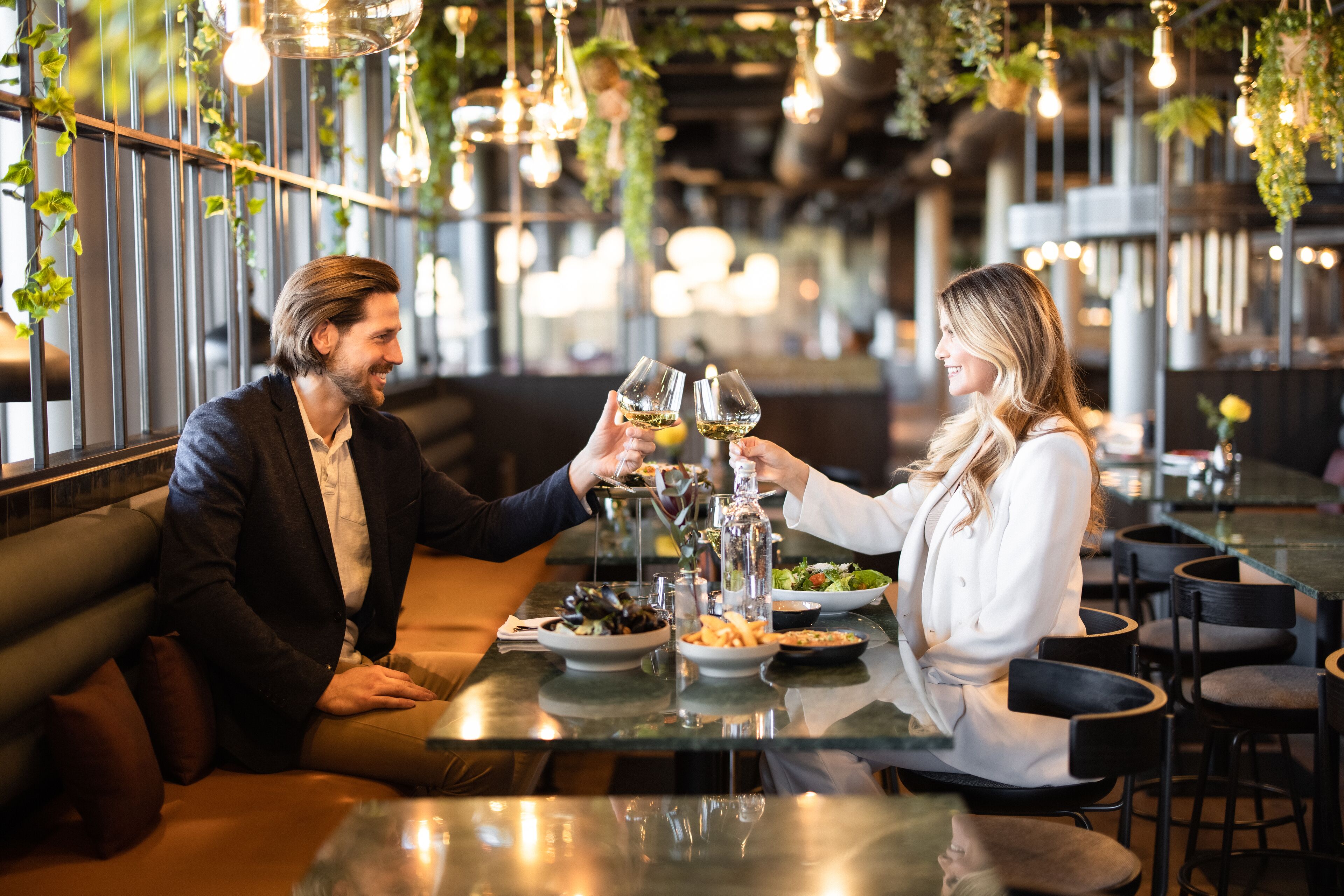 Um casal brindando com taças de vinho em um restaurante com decoração moderna, incluindo luminárias pendentes e uma mesa de vidro.