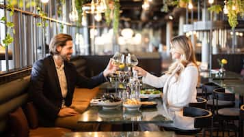 Um casal brindando com taças de vinho em um restaurante com decoração moderna, incluindo luminárias pendentes e uma mesa de vidro.