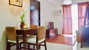 Uma sala de jantar com uma mesa de madeira, cadeiras de vime, um vaso com uma planta, uma televisão sobre um suporte e uma porta.