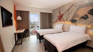 Quarto de hotel com duas camas, uma secretária e uma janela grande com cortinas.