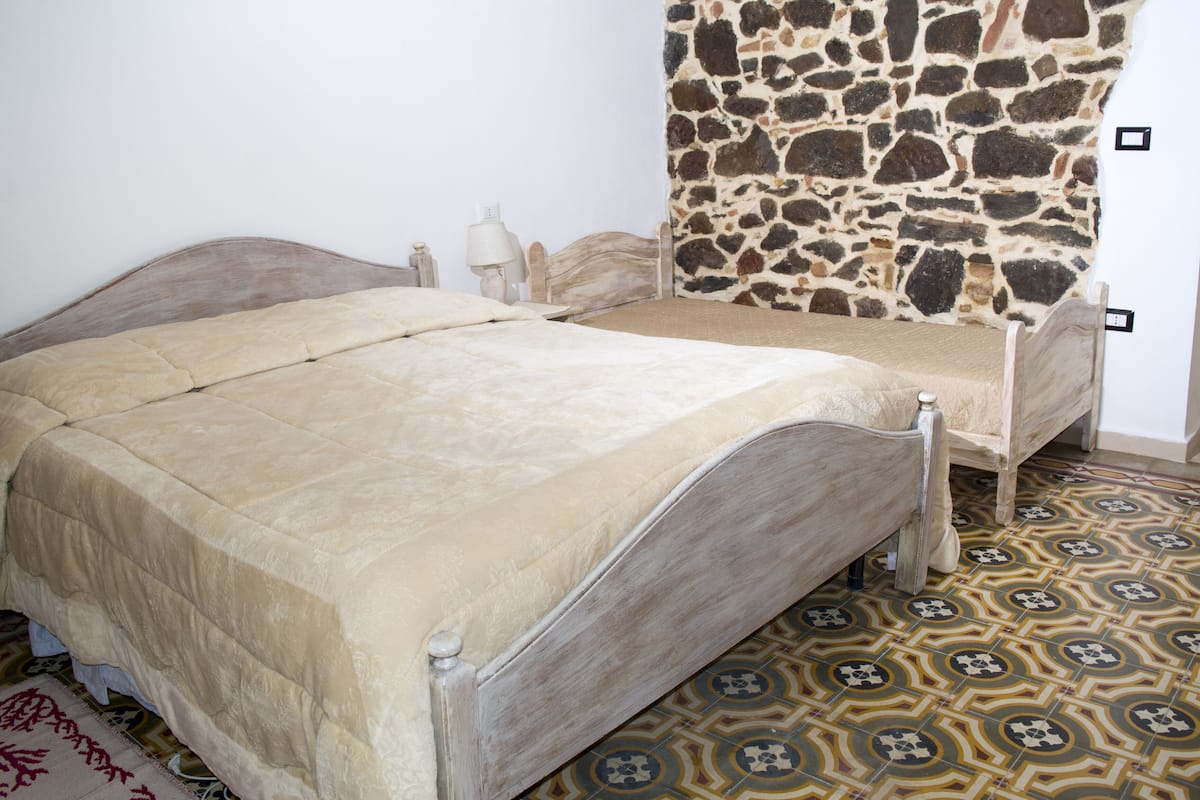 Quarto com cama, mesa de cabeceira, luminária e parede de pedra.