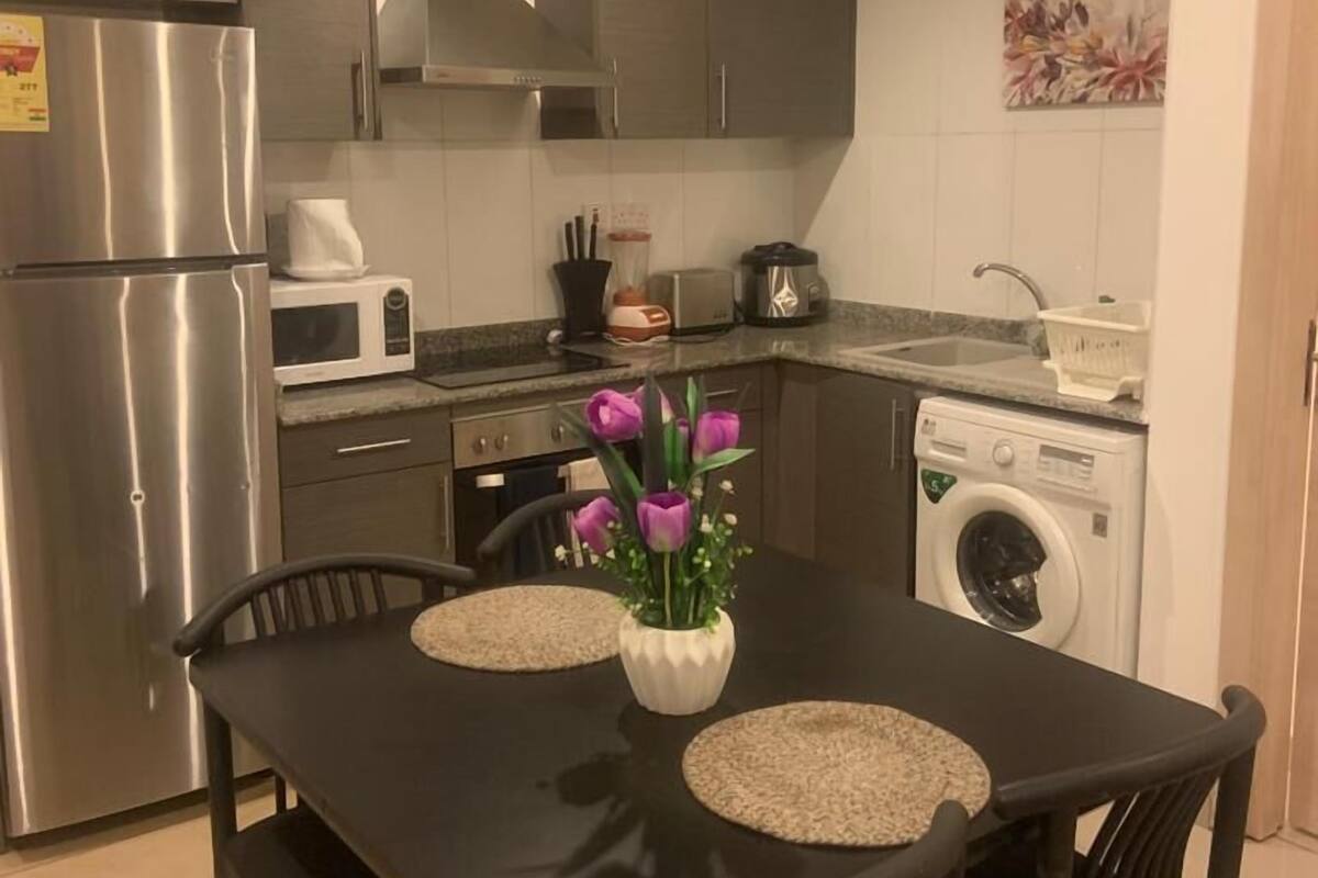 Cozinha compacta com geladeira, micro-ondas, torradeira e máquina de lavar. Há uma mesa de jantar com cadeiras e um vaso de flores.