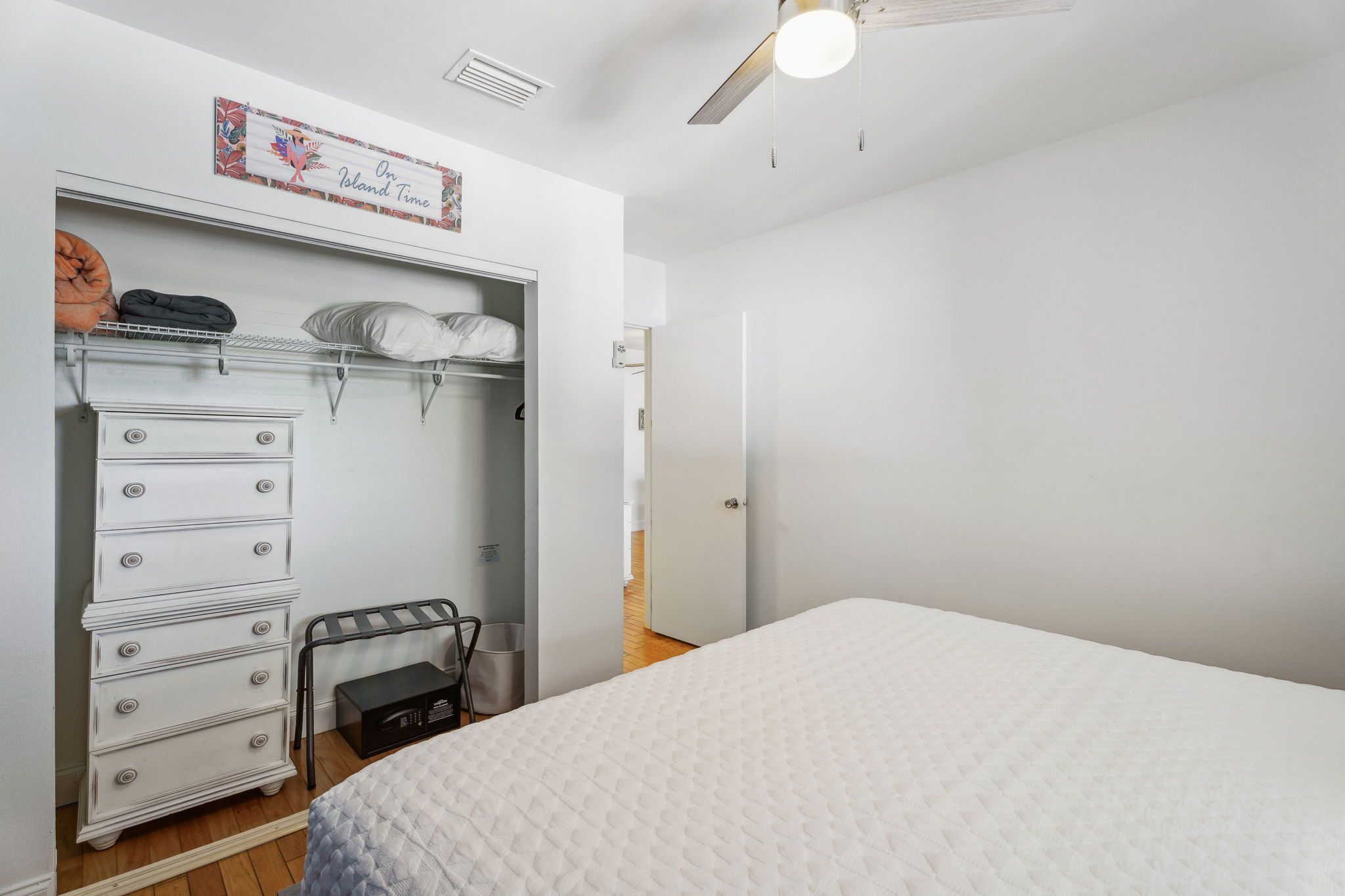 Quarto com cama, cômoda branca, armário com roupas penduradas e ventilador de teto.
