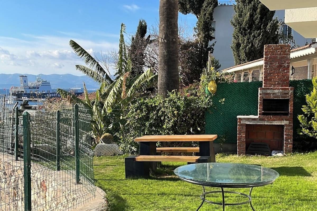 Um pátio com mesa de vidro e banco, rodeado por vegetação e vista para o mar.