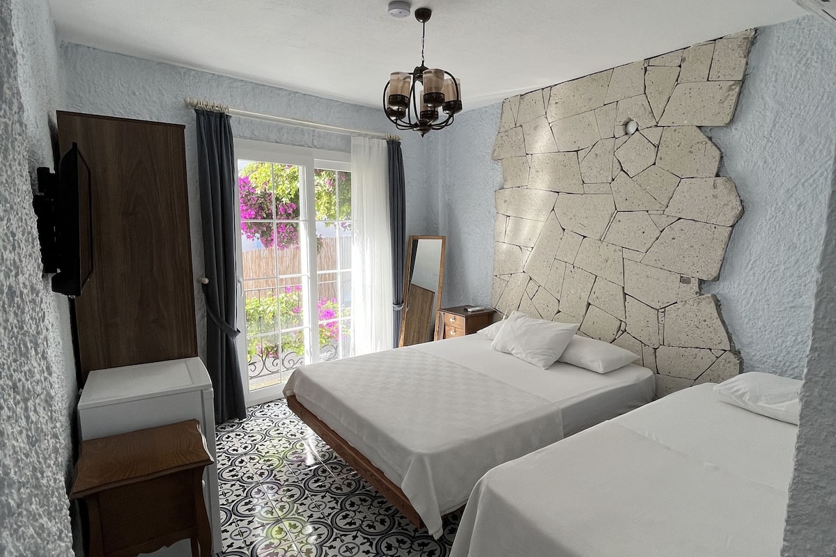 Quarto de hotel com duas camas, um banco de madeira, uma TV e uma janela grande com cortinas.