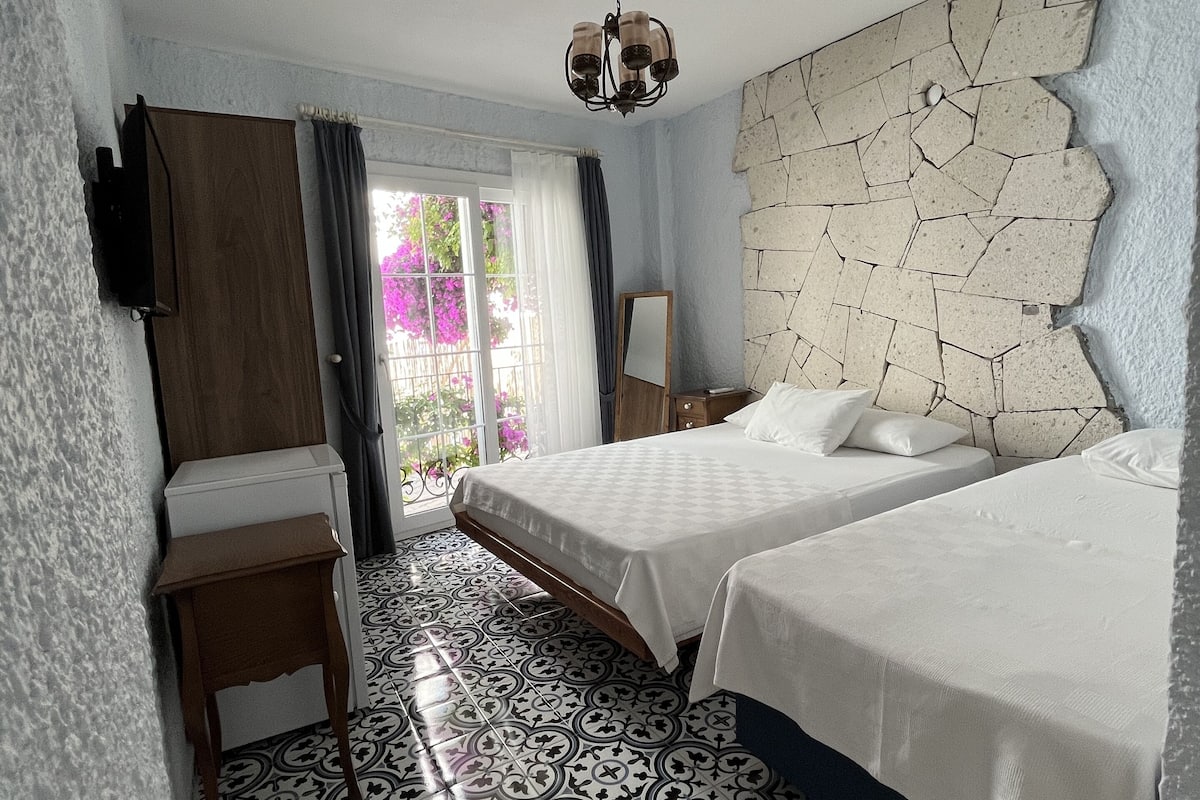 Quarto de hotel com duas camas, um guarda-roupa de madeira, uma mesa pequena, uma TV fixada na parede e um chão estampado.