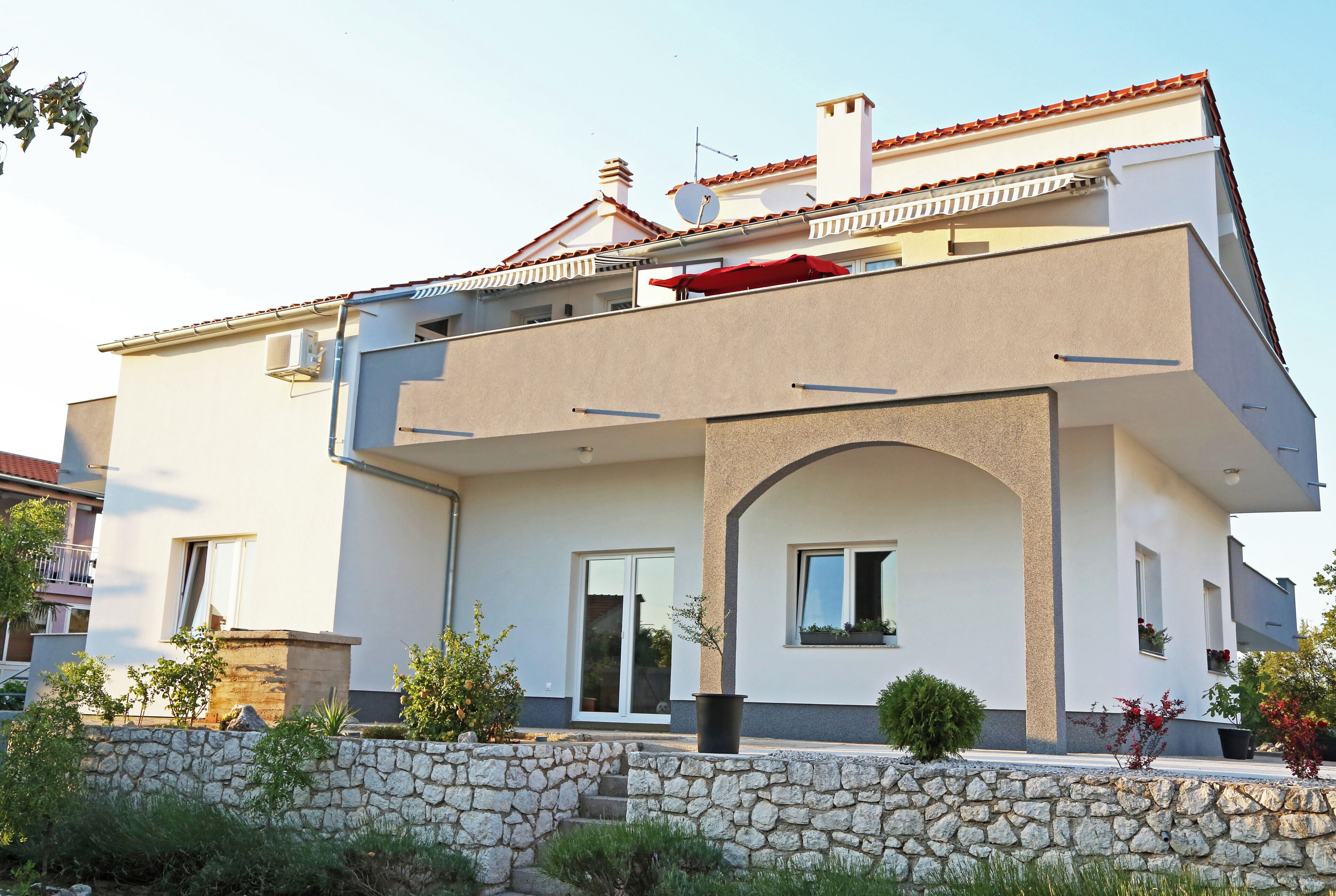 Uma casa moderna de dois andares com uma parede de pedra em frente, uma varanda com guarda-sol vermelho e uma antena parabólica no telhado.