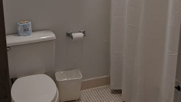 Um banheiro pequeno com vaso sanitário, um porta-toalhas com duas toalhas, um rolo de papel higiênico, uma cortina de chuveiro e uma lixeira.