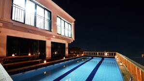 Vista noturna de um hotel com uma grande piscina ao ar livre, várias janelas e um guarda-corpo de varanda.