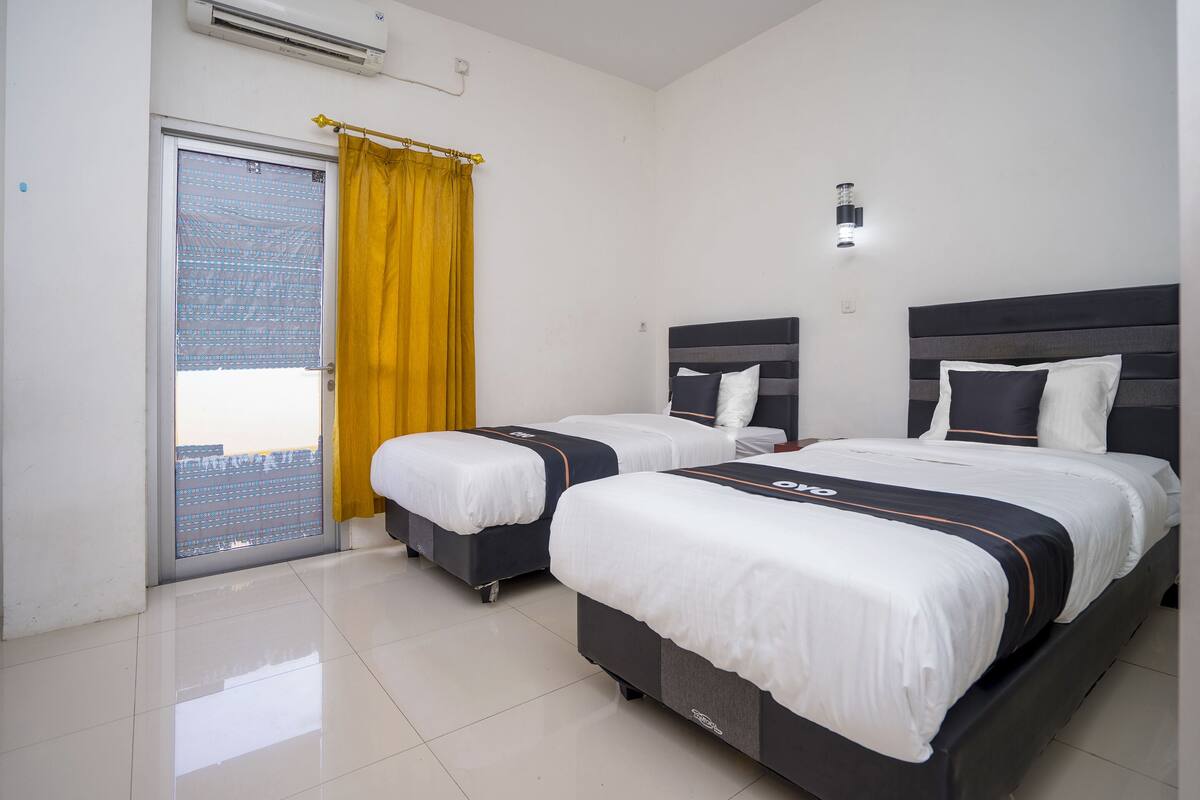 Quarto de hotel com duas camas, uma porta de correr de vidro e um ponto de luz fixado na parede.