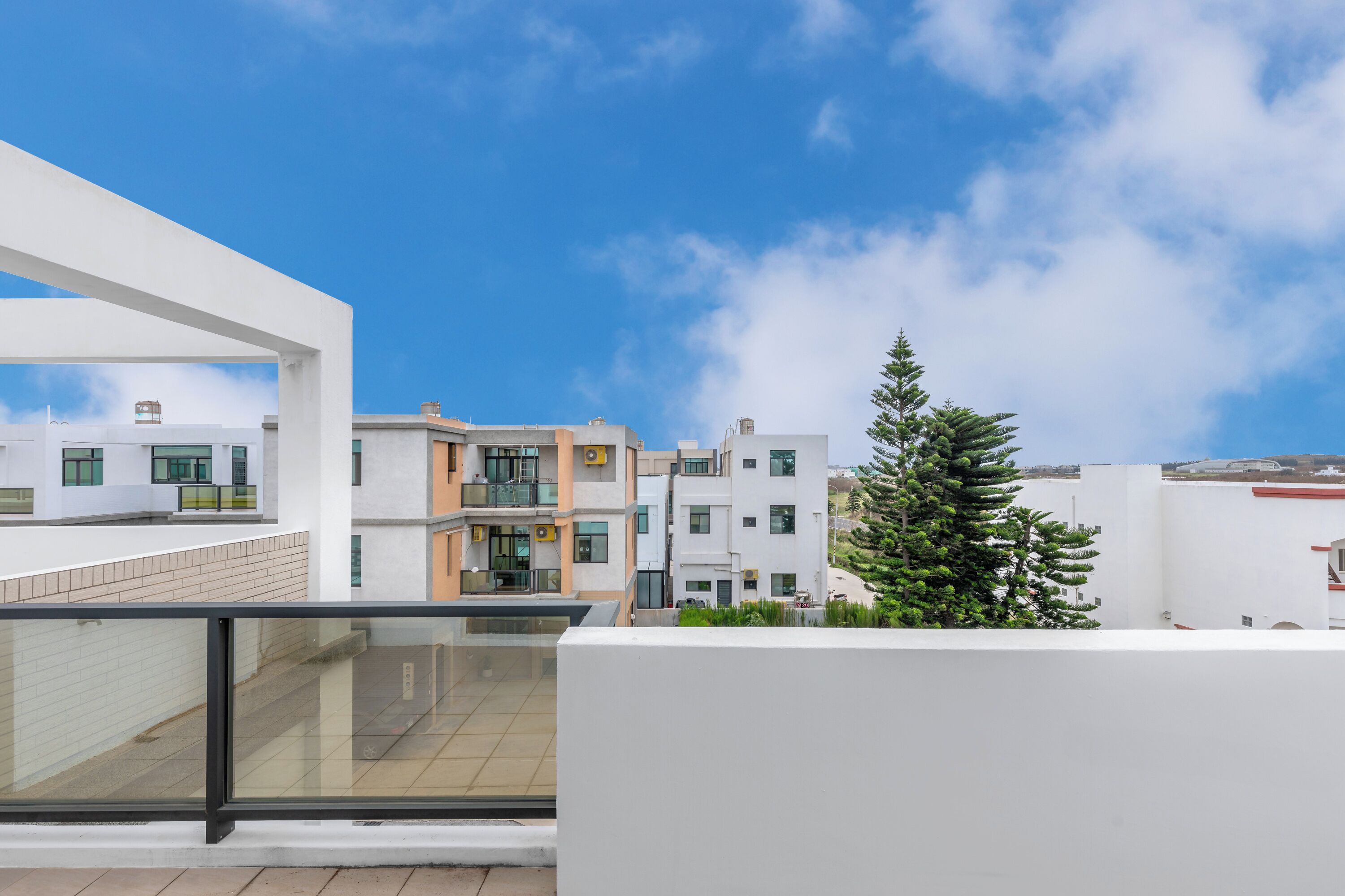 Um edifício residencial moderno com varanda, céu claro e uma árvore em primeiro plano.