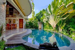 Piscina privativa com deck de madeira, cercada por vegetação exuberante e uma parede de pedra.
