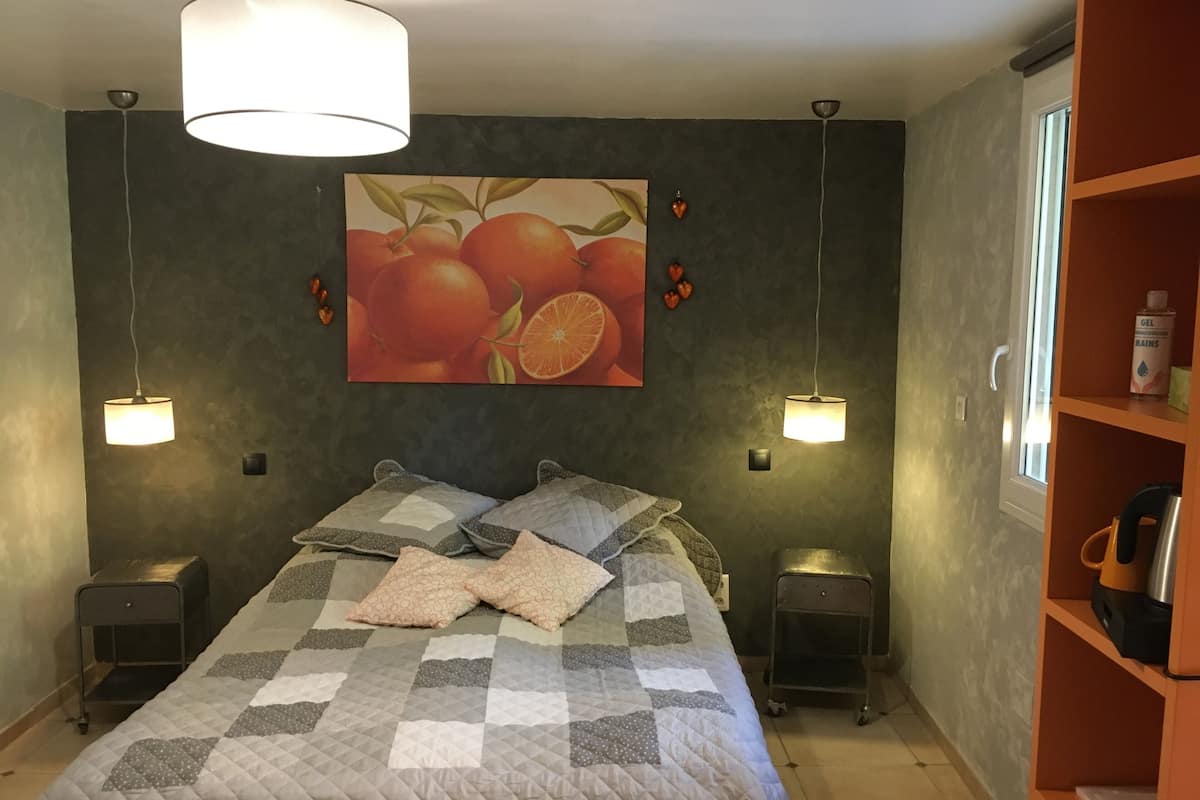 Quarto com cama, duas mesinhas de cabeceira, um criado-mudo, um quadro na parede com laranjas e uma estante com chaleira e garrafa.