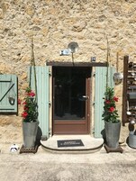 Um edifício rústico com porta de madeira e entrada com persianas, dois vasos com flores vermelhas, e uma placa indicando o preço de 15 euros.