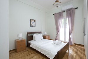 Quarto bem arrumado com uma cama de madeira, uma mesinha de cabeceira com abajur, um pequeno móvel com gavetas e uma janela com cortinas.