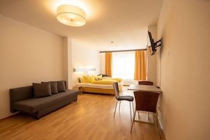 Quarto de hotel moderno com sofá, cama com roupa de cama amarela, escrivaninha com cadeira e uma janela grande com cortinas.