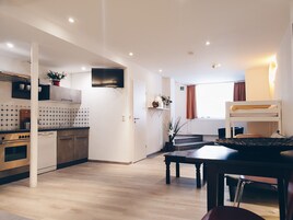 Uma cozinha moderna com armários brancos, bancada de madeira e forno embutido. Há uma área de jantar com mesa e cadeiras, e uma cama beliche ao fundo.