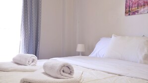 Cama bem arrumada com lençóis brancos e toalhas dobradas, mesa de cabeceira branca com luminária, e um quadro de um caminho de cerejeiras na parede.