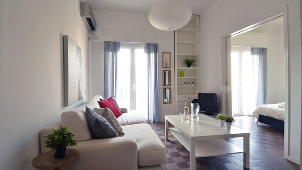Uma sala de estar moderna com um sofá, uma mesa de jantar e uma televisão. Há uma estante de livros e uma janela com cortinas.