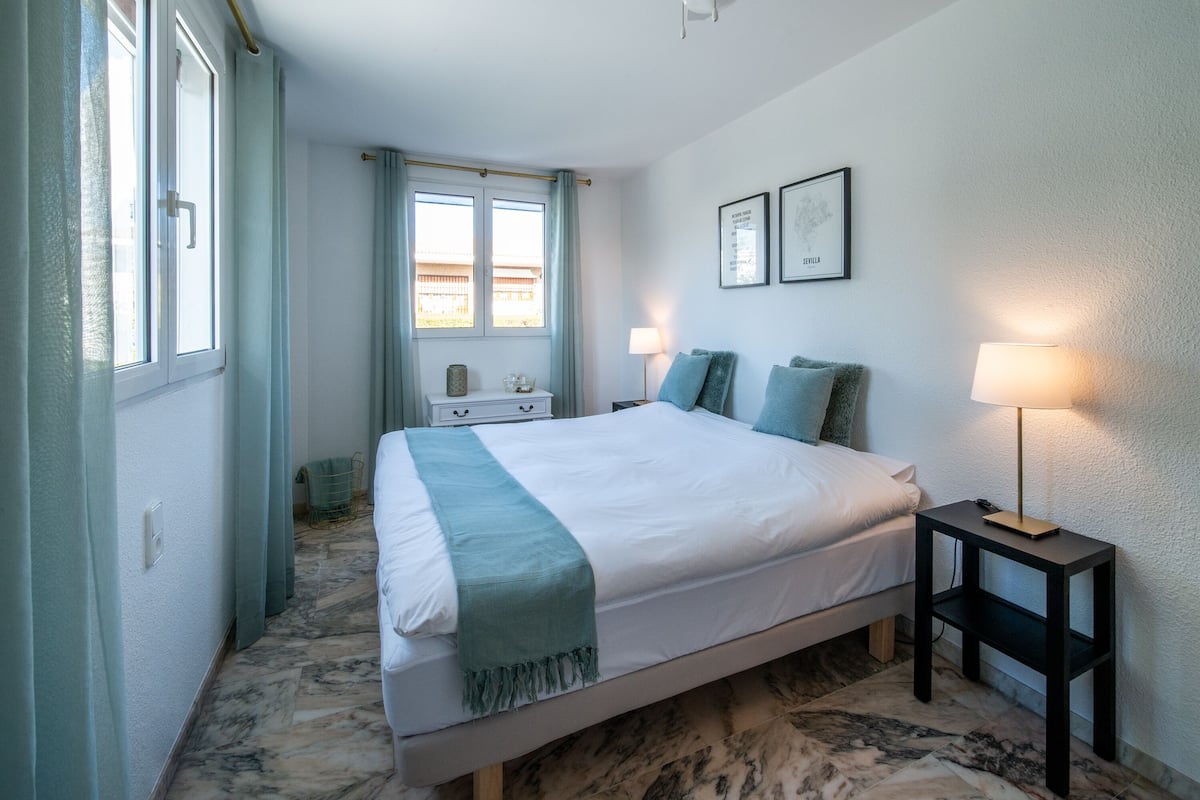 Quarto com cama, duas luminárias de cabeceira, mesa de cabeceira, janela com cortinas e parede com duas molduras.