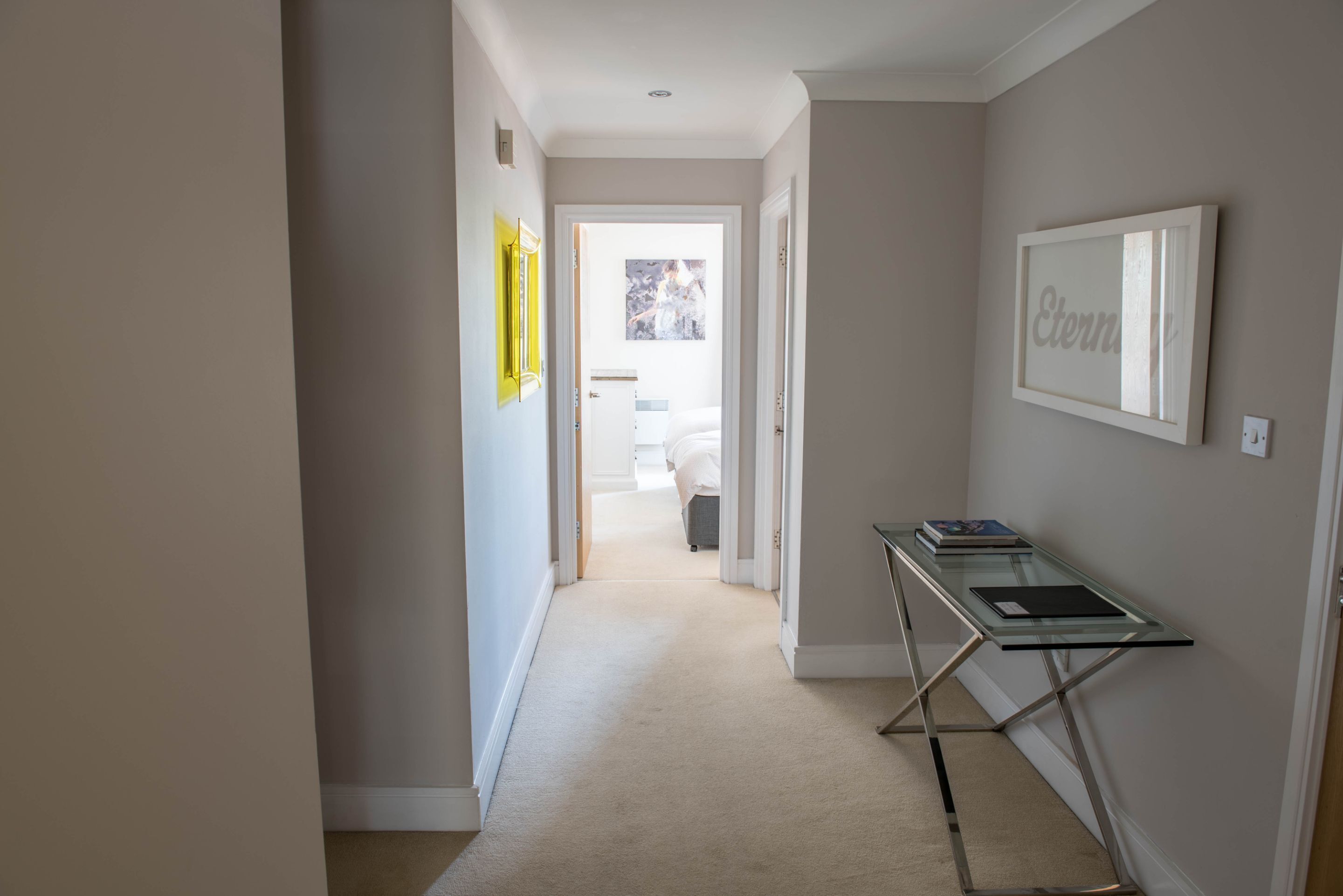 Um corredor com uma mesa de vidro, um quadro na parede e uma porta que dá para um quarto.