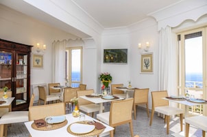 Uma sala de jantar com mesas e cadeiras brancas, um armário de madeira e vista para o mar.