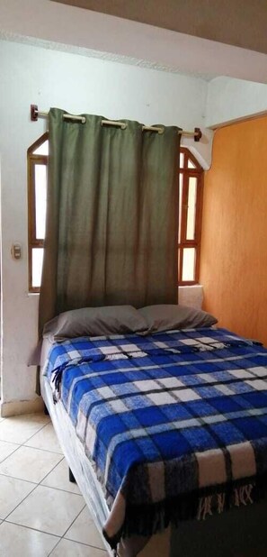 Uma cama de solteiro com colcha azul e branca xadrez, cortina verde e janela com molduras de madeira.
