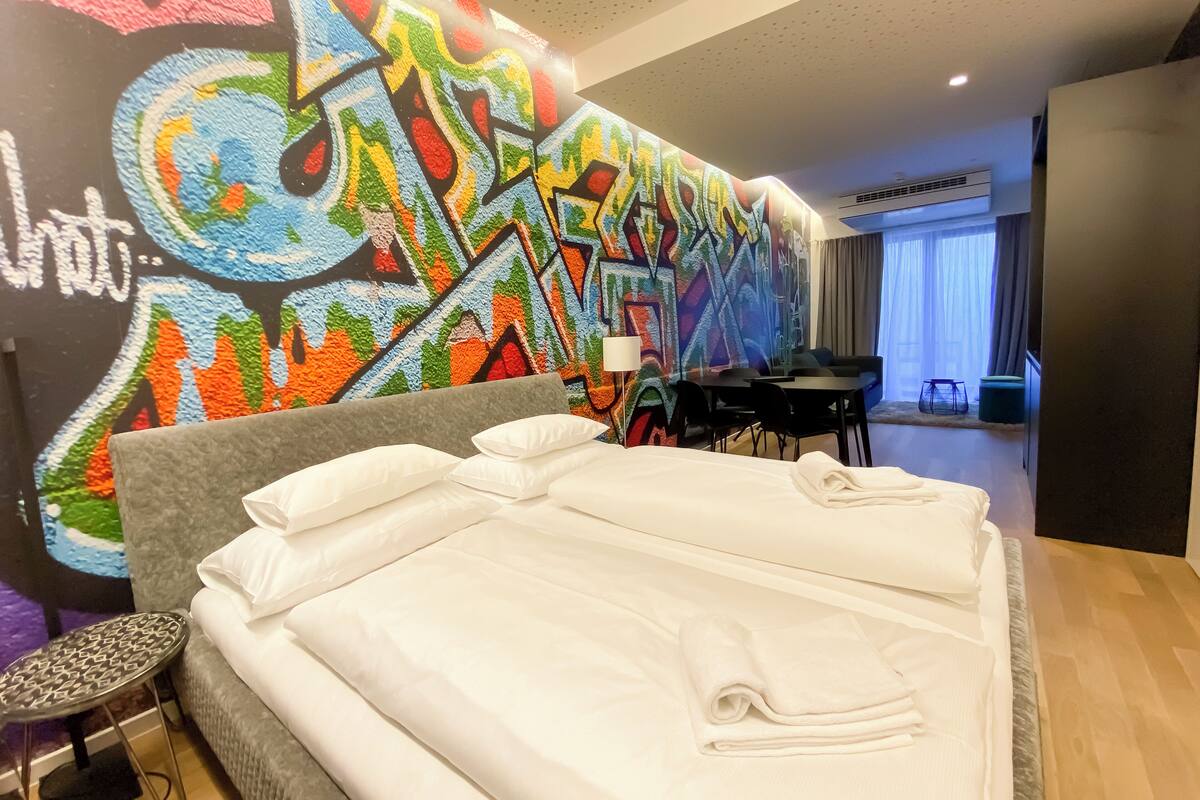 Quarto de hotel com uma cama grande, uma escrivaninha com cadeira, uma mesinha e um mural colorido na parede.