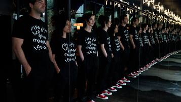 Uma fila de pessoas em frente a uma superfície refletora com texto e uma fileira de sapatos vermelhos.