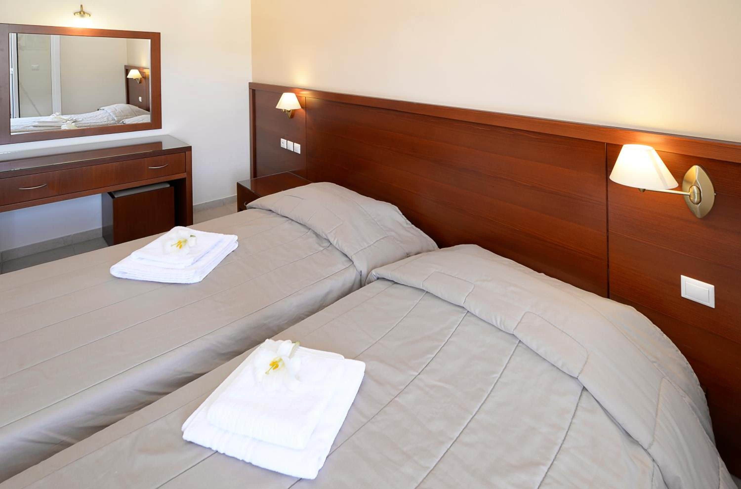 Quarto de hotel com duas camas, cabeceira de madeira, abajures, um espelho e uma pequena escrivaninha com lâmpada.