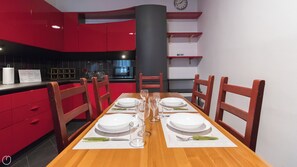 Uma sala de jantar com uma mesa de madeira posta para quatro pessoas, cadeiras vermelhas e uma cozinha ao fundo.
