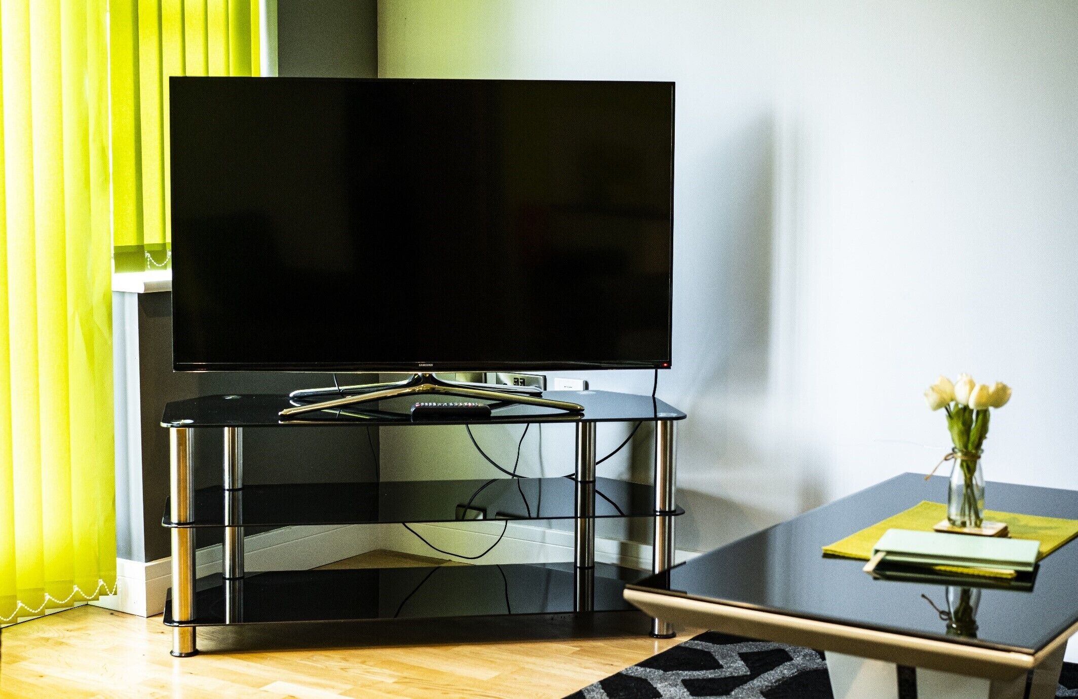 Uma sala de estar moderna com uma televisão de tela plana em um suporte preto, uma mesinha com um vaso de flores e uma cortina amarelo vibrante.
