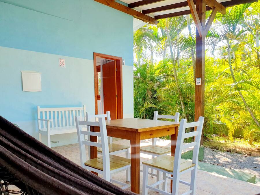 Quarto com mesa de madeira, cadeiras brancas, uma rede e porta que dá para o exterior.