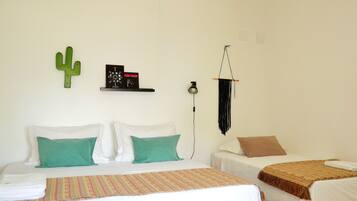 Quarto de hotel com cama, duas mesas de cabeceira, prateleira na parede com objetos decorativos, luminária pendente e cacto decorativo.
