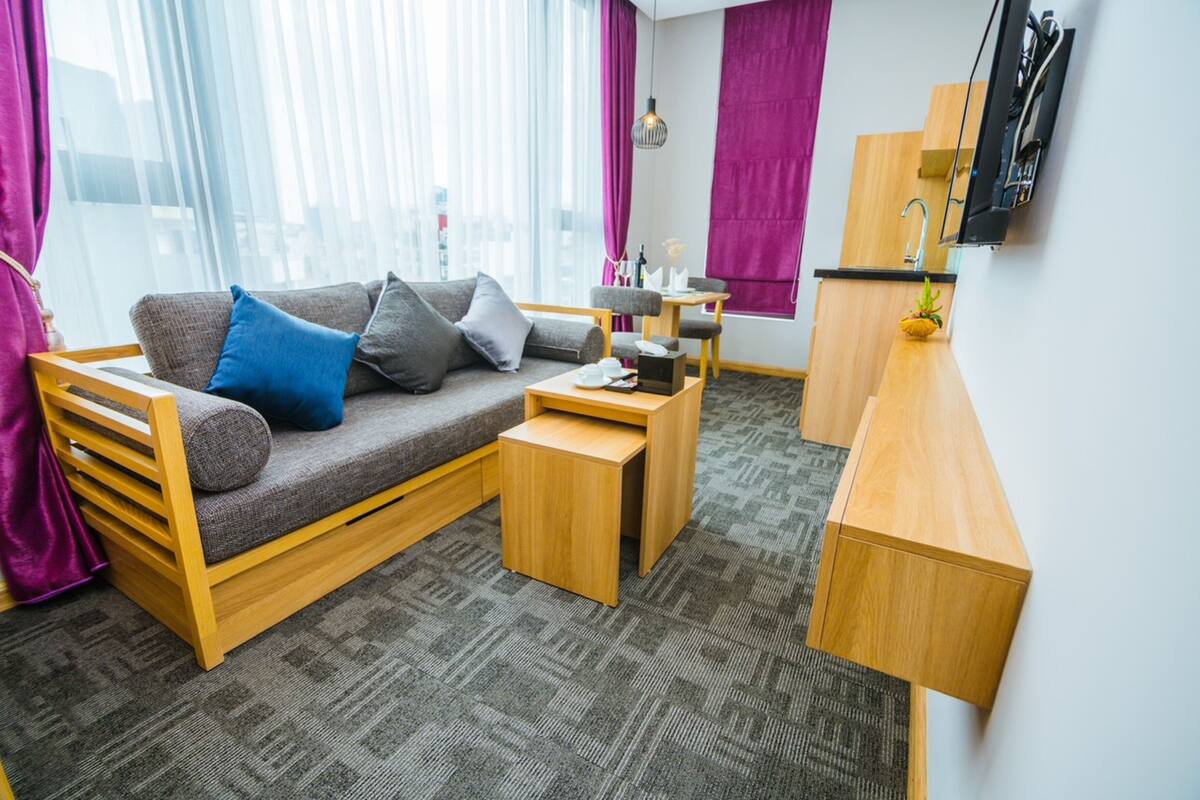 Quarto de hotel moderno com sofá, uma pequena mesa de madeira, TV na parede e vista da cidade por grandes janelas.