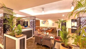 Um hall de hotel espaçoso com área de refeições central, várias opções de assento e plantas decorativas.