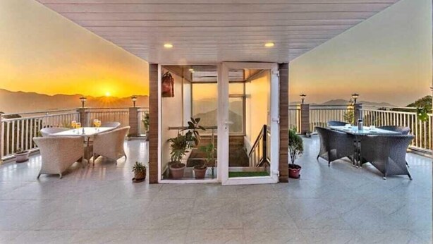 Varanda com mobiliário de vime e plantas em vasos, com vista para um pôr do sol e montanhas ao longe.