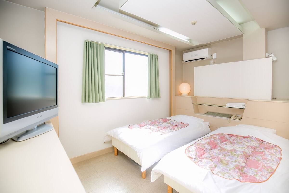 Quarto de hotel com duas camas de solteiro, televisão de tela plana, mesa de cabeceira, janela com cortinas, ar-condicionado e luminária de cabeceira.