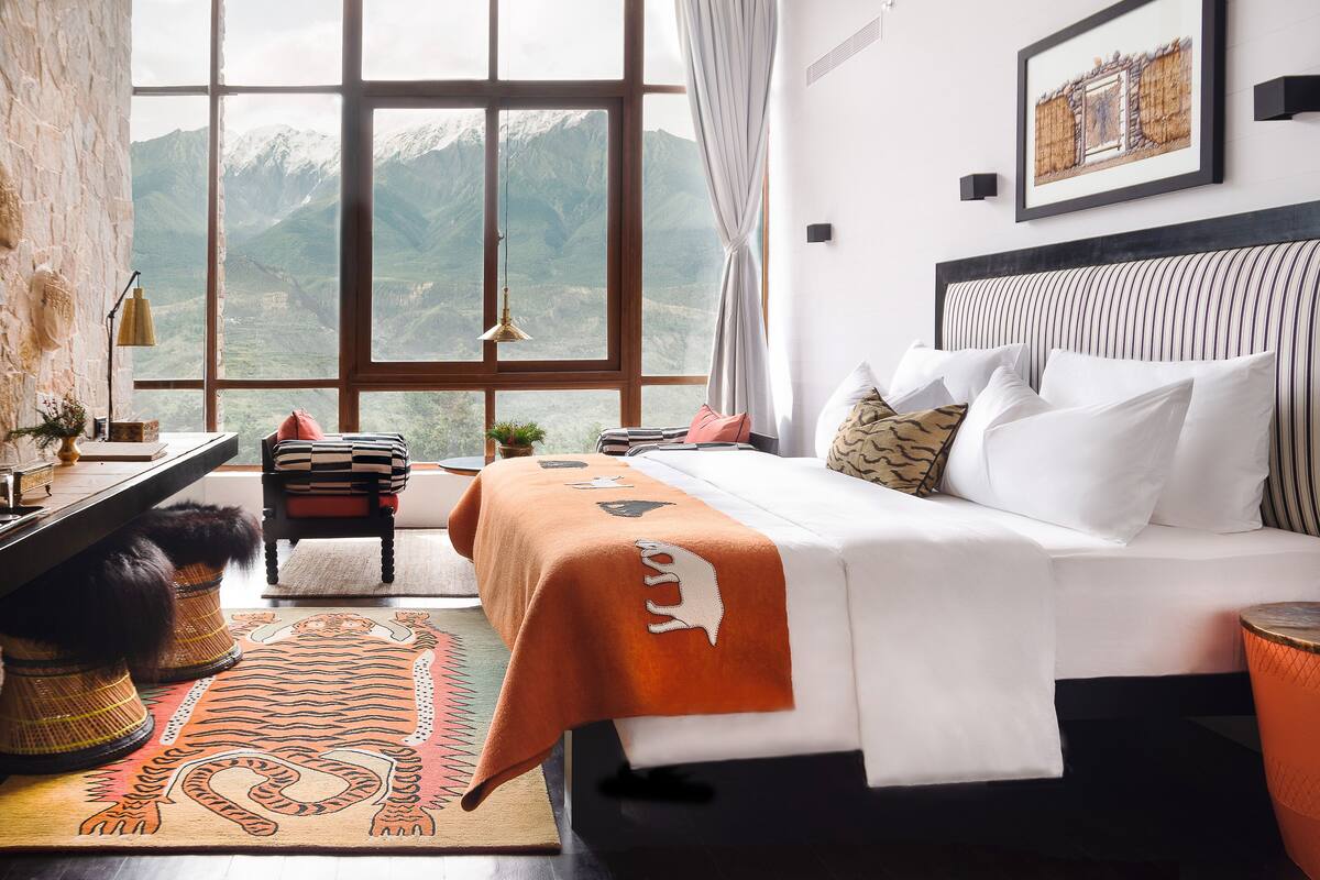 Quarto com uma janela ampla, cama com roupa de cama listrada, escrivaninha com luminária e vista para montanhas.