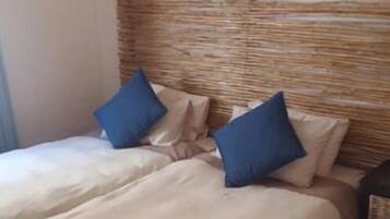 Uma cama de casal com roupa de cama branca e almofadas azuis, cabeceira de vime e um quadro na parede.