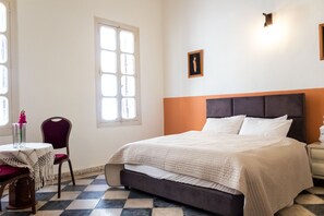 Quarto de hotel com piso xadrez, uma cama com cabeceira, uma mesa com um vaso e uma cadeira.