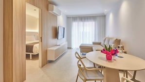 Quarto de hotel moderno com área de refeições, sofá, TV fixada na parede e uma grande janela com cortinas.