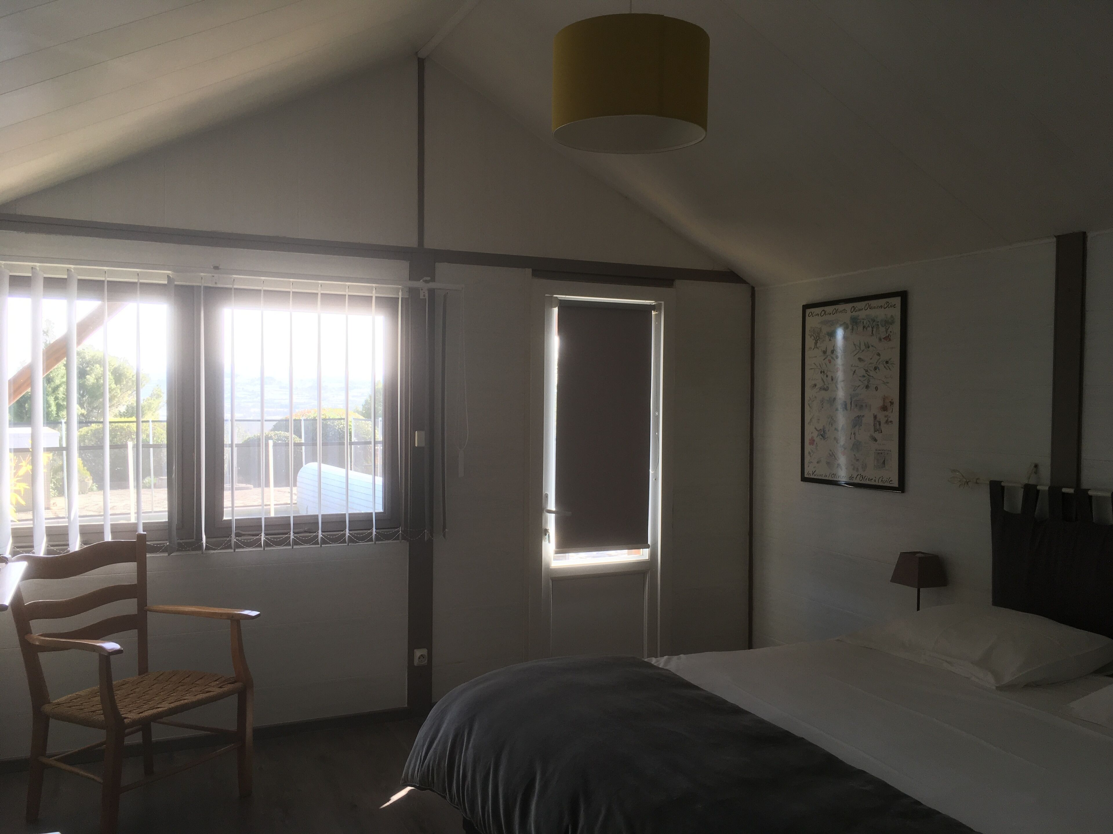Quarto com cama, uma cadeira, janela com persianas, porta e luminária fixada na parede.