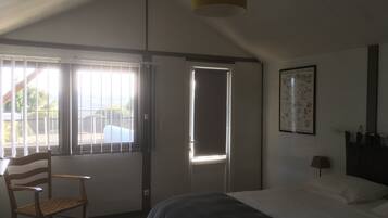 Quarto com cama, uma cadeira, janela com persianas, porta e luminária fixada na parede.