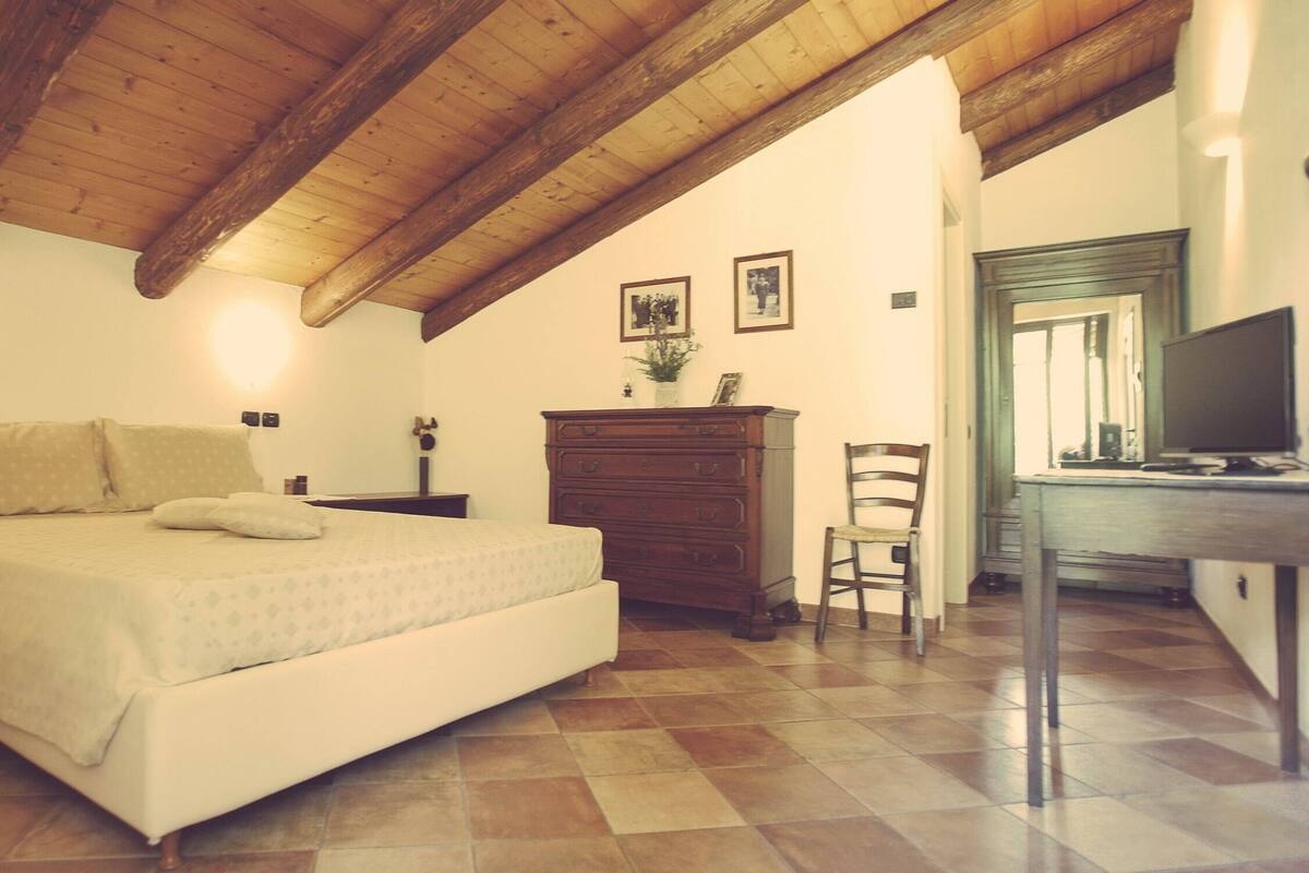 Quarto com cama, vigas de madeira no teto, cômoda, cadeira e televisão.