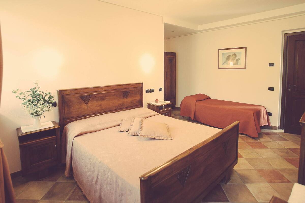 Quarto de hotel com cama de madeira, mesa de cabeceira com abajur, pequena mesa com um vaso de flores e porta que dá para outro quarto.