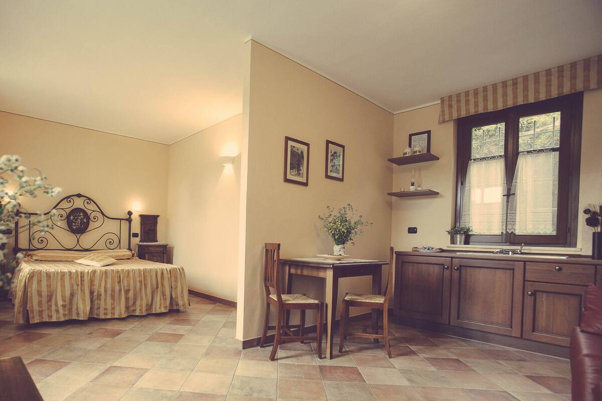 Quarto com cama, mesa de cabeceira e abajur; pequena área de jantar com mesa e cadeira; cozinha com armários e janela; e banheiro com vaso sanitário e pia.