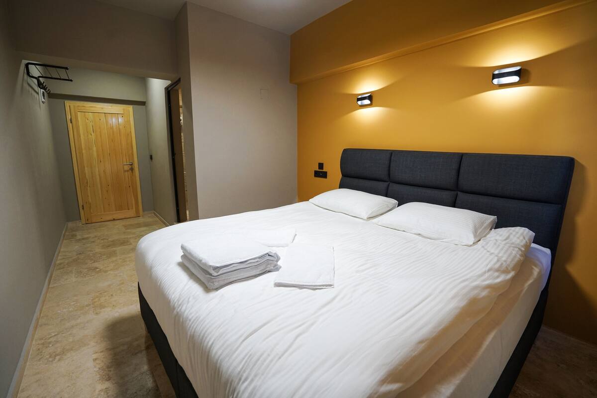 Cama bem arrumada com lençóis brancos e uma toalha dobrada, porta de madeira ao fundo e duas luzes fixadas no cabeceira da cama.